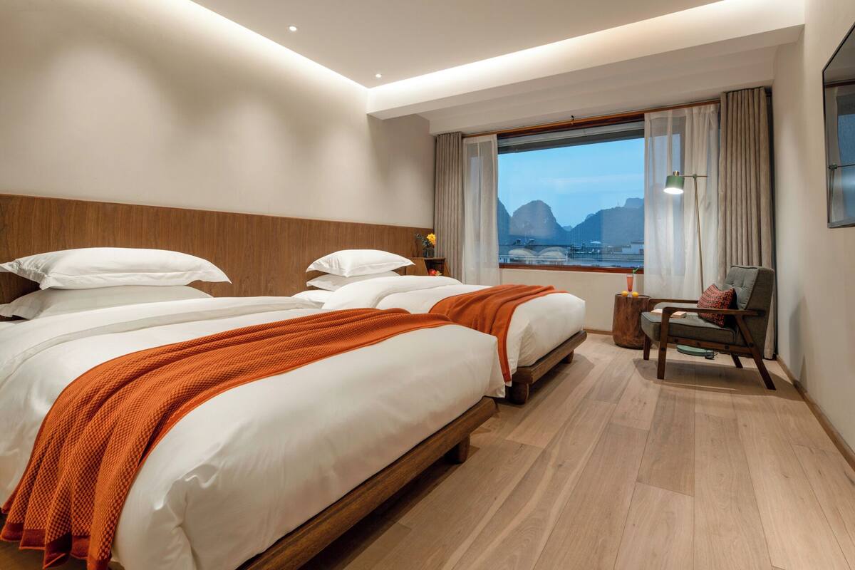 Quarto de hotel com duas camas, cabeceira de madeira, vista para montanhas, escrivaninha com cadeira e uma luminária.
