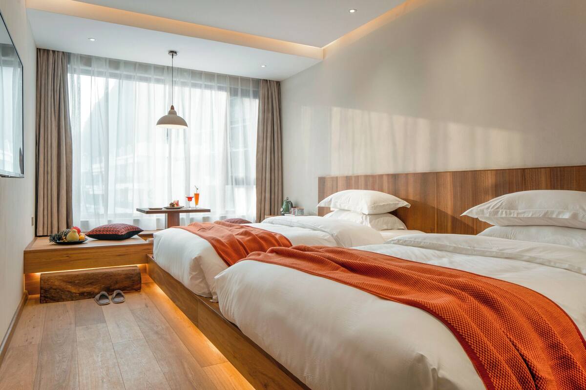 Quarto de hotel moderno com uma cama grande, uma mesa de cabeceira com abajur e vista para a cidade através de cortinas leves.