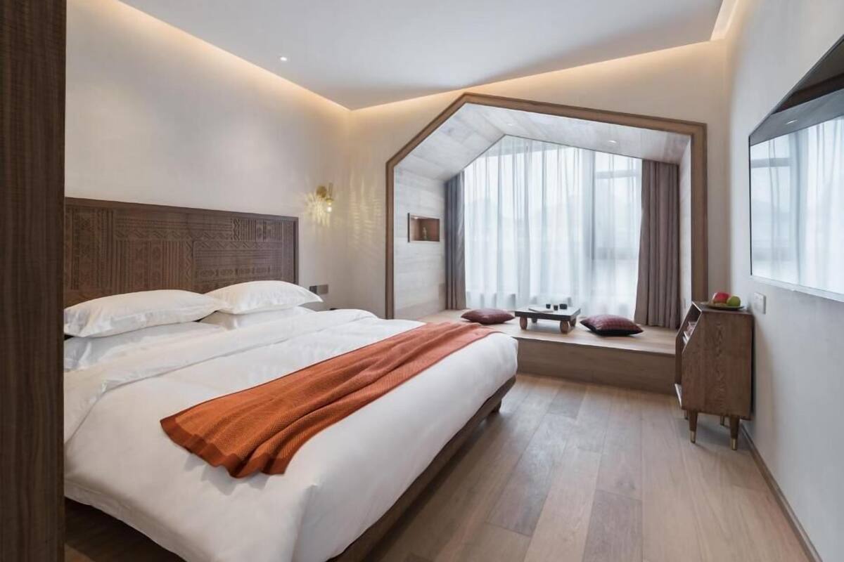 Quarto de hotel moderno com uma cama grande, um pequeno espaço de estar junto à janela e uma mesinha de cabeceira de madeira.