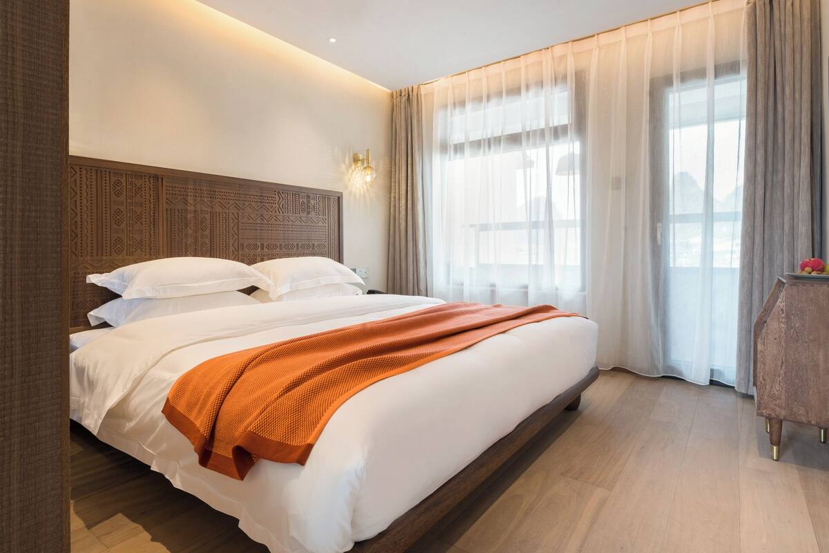 Quarto de hotel moderno com uma cama grande, cabeceira com design estampado, luminária fixada na parede e vista através de cortinas leves.