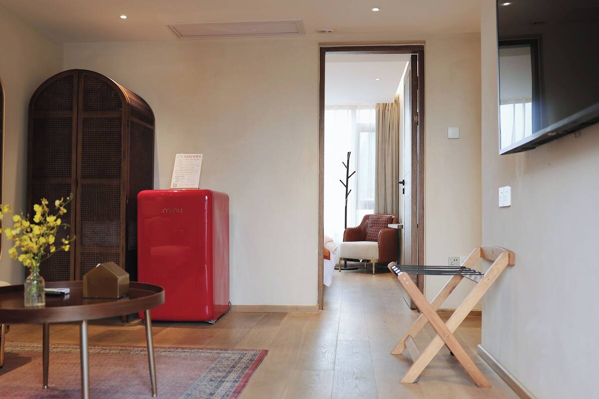 Uma sala de estar moderna com um mini-frigorífico vermelho, um porta-roupas de madeira, uma TV fixada na parede e vista para outro cômodo com uma cadeira e um porta-roupas.