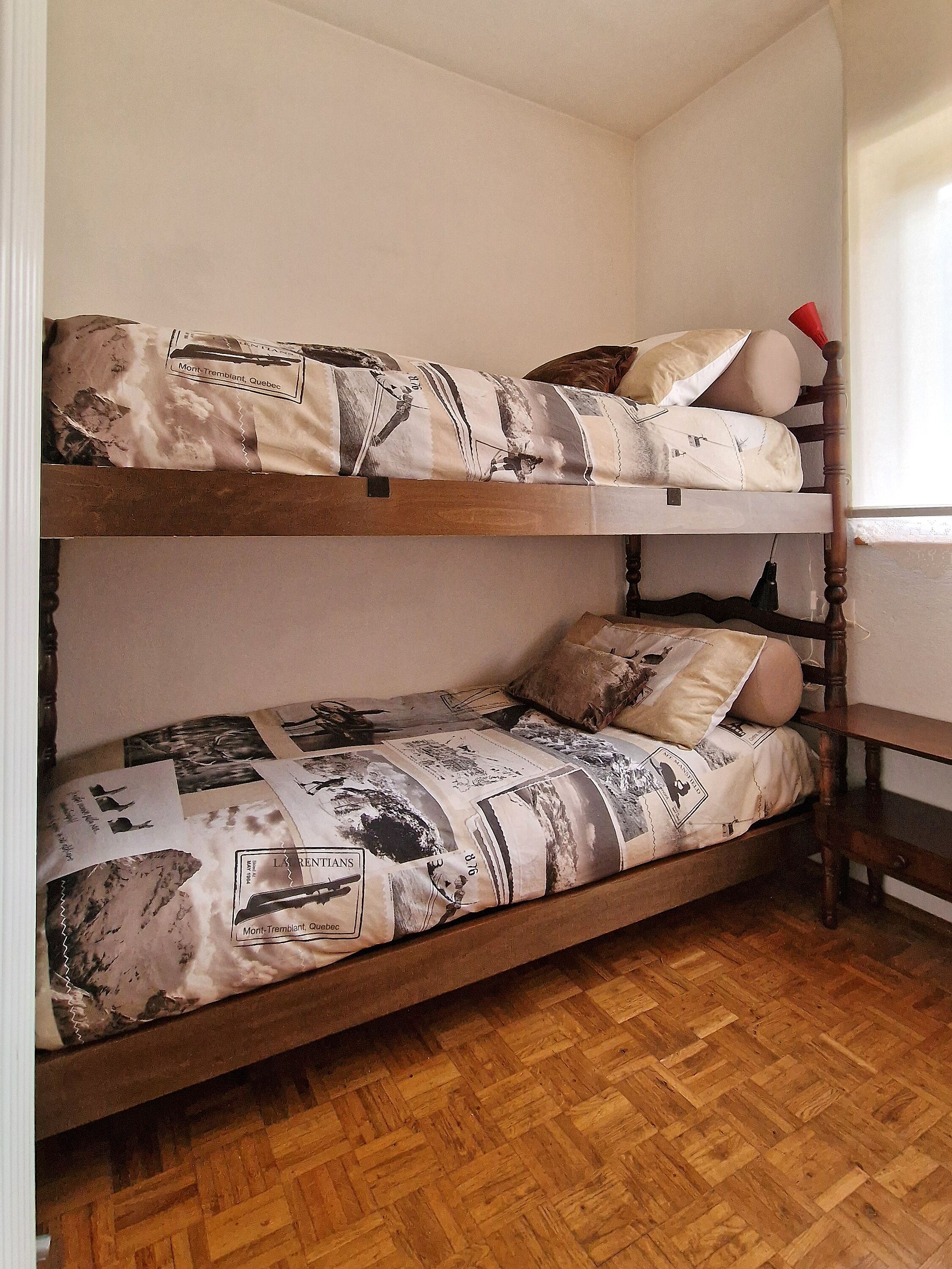 Uma cama beliche com colcha estampada e almofadas, piso de madeira e uma janela com cortinas.