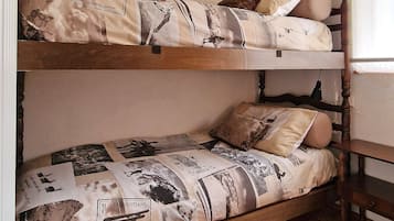 Uma cama beliche com colcha estampada e almofadas, piso de madeira e uma janela com cortinas.
