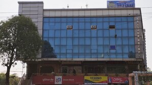 Um edifício com fachada de vidro azul, uma escadaria à esquerda e diversas lojas, incluindo um banco e uma loja de eletrônicos.