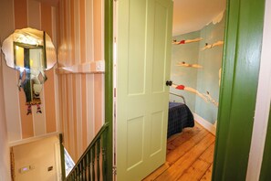 Quarto com piso de madeira, cama com manta azul, porta verde e parede com padrão listrado.