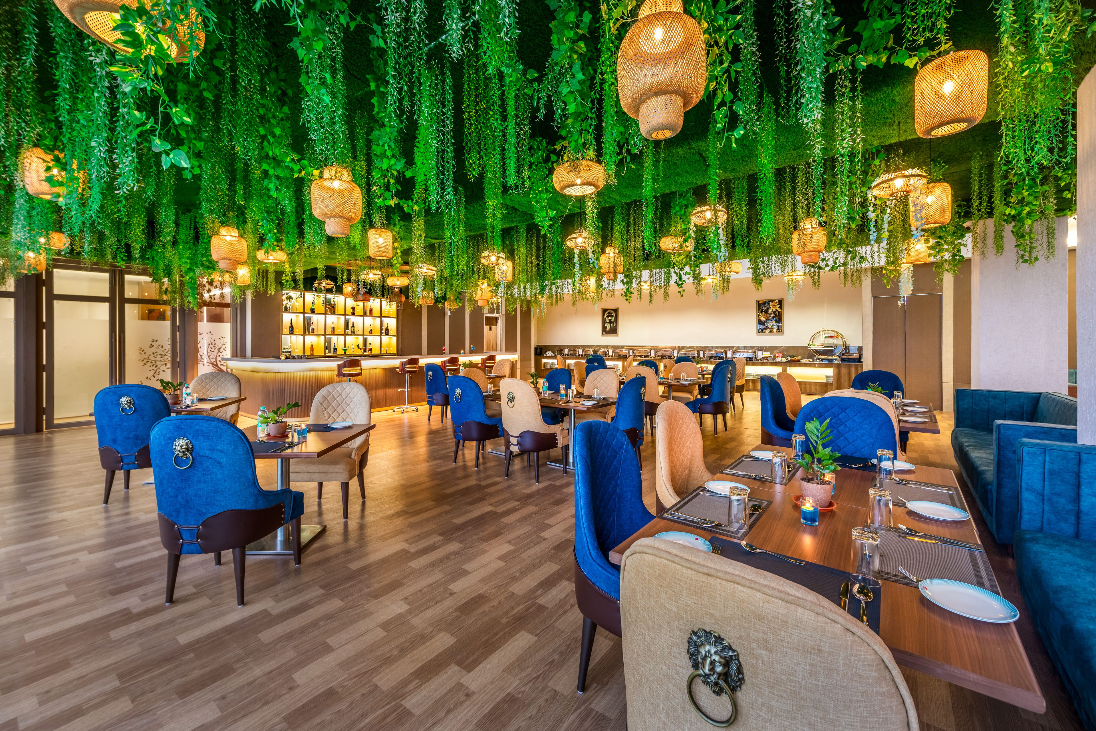 Restaurante com plantas pendentes e luminárias de vime, piso de madeira e cadeiras estofadas em azul.