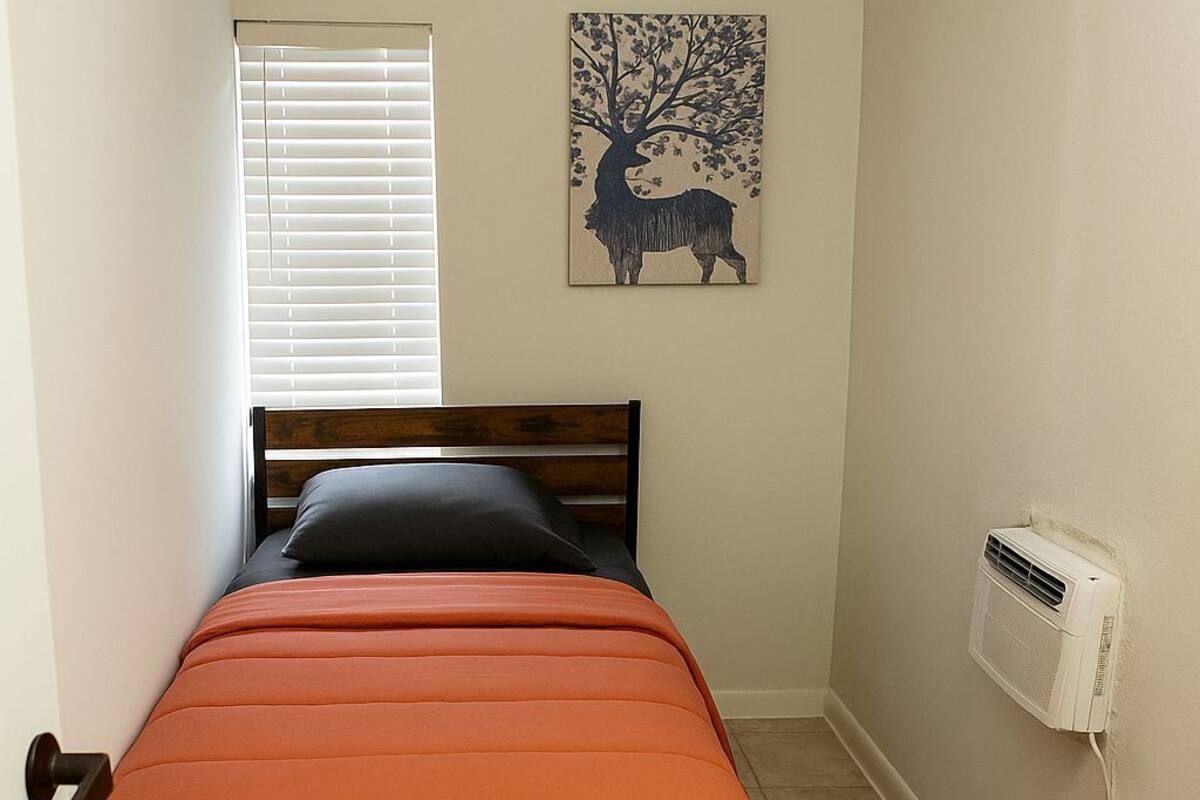 Quarto com cama, janela com persianas, ar condicionado fixado na parede e uma ilustração com silhueta de árvore.