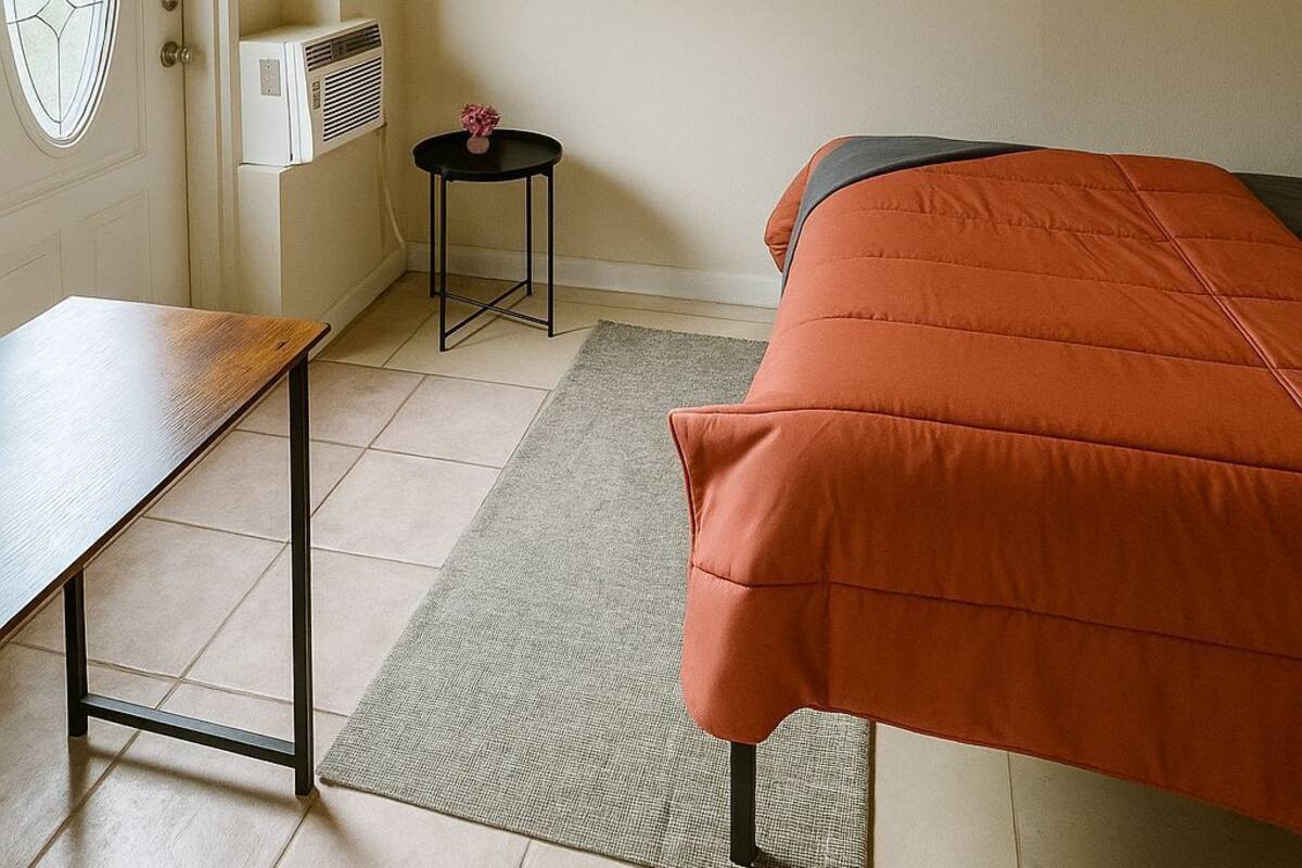 Quarto com cama, ventilador de teto, aquecedor, mesa pequena e porta com janela.