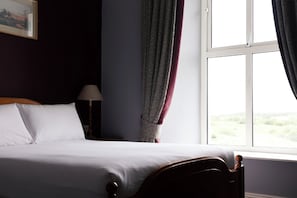 Uma cama bem arrumada com lençóis brancos, cabeceira de madeira, luminária de cabeceira e uma janela com cortinas.