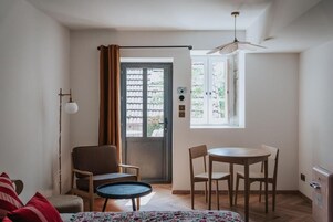 Quarto com cama, cadeira, mesa pequena, mesa de jantar, ventilador de teto e janela com cortinas.