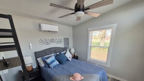 Um quarto com cama, ventilador de teto, ar condicionado, janela com vista e porta com puxador.