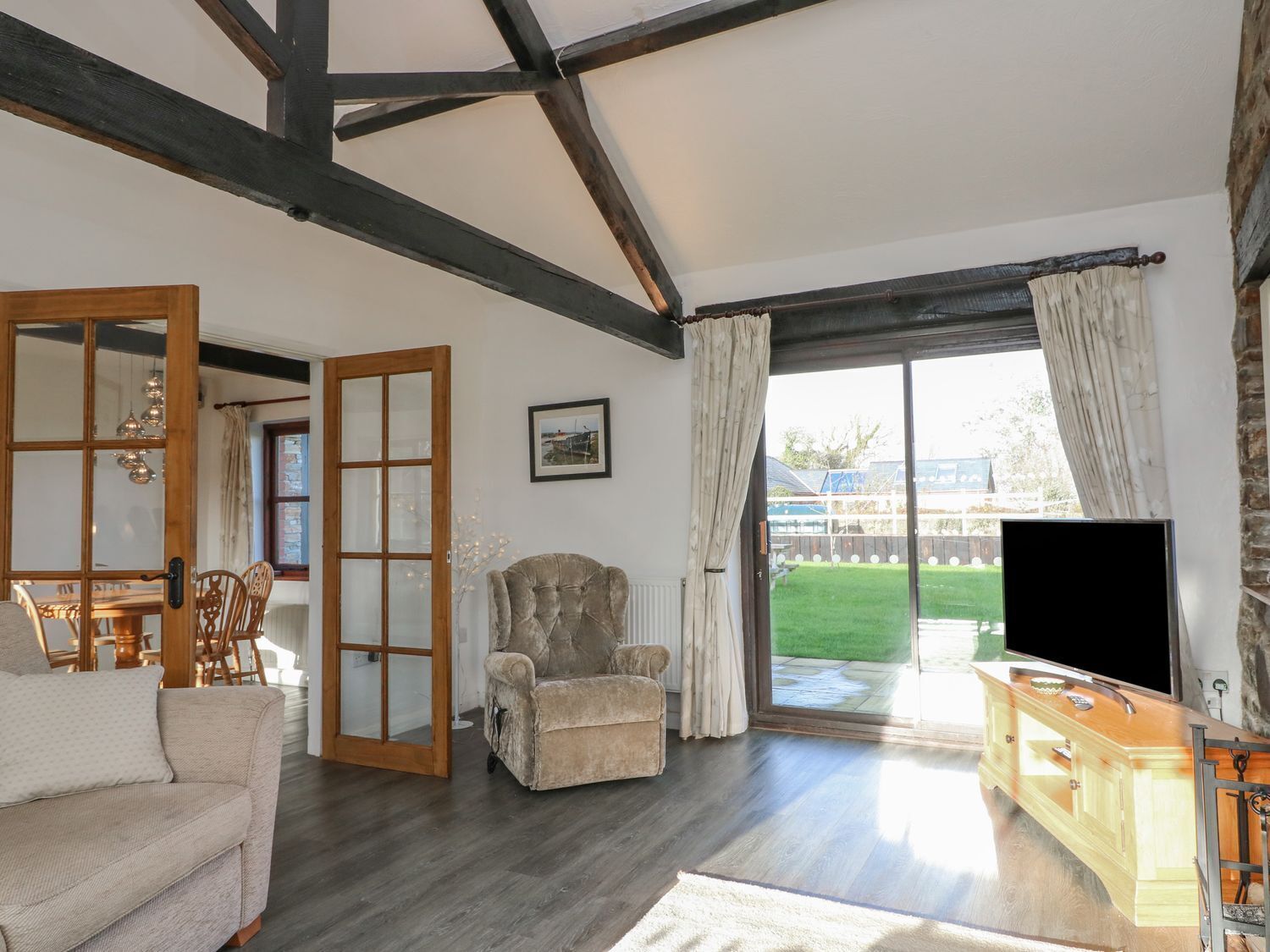 Uma sala de estar ampla com teto de madeira, uma grande porta de correr de vidro que dá para um jardim, uma mesa de jantar de madeira e uma TV em um móvel de madeira.