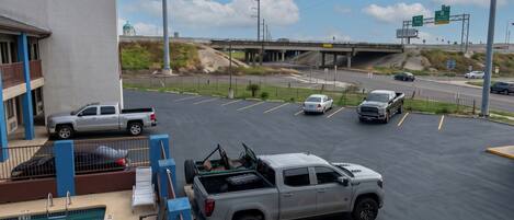Um parque de estacionamento com piscina, um edifício com fachada azul e branca, e uma passarela rodoviária ao fundo.