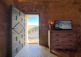 Uma porta aberta que dá para uma varanda com vista, um aparador de madeira com uma televisão, e um vaso com flores.