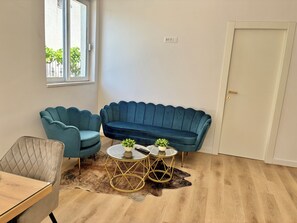 Um quarto com um sofá azul, uma mesa de centro redonda, uma mesinha com uma planta, uma cadeira e uma porta.