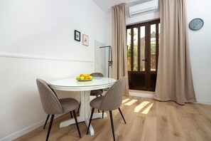 Uma área de refeições com mesa de vidro, duas cadeiras, um cesto de frutas, relógio fixado na parede e uma porta de correr de vidro.