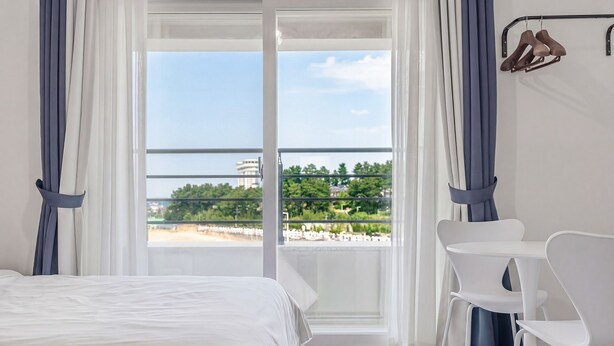 Quarto de hotel com cama, varanda com cortinas e vista para a praia, além de uma cadeira.