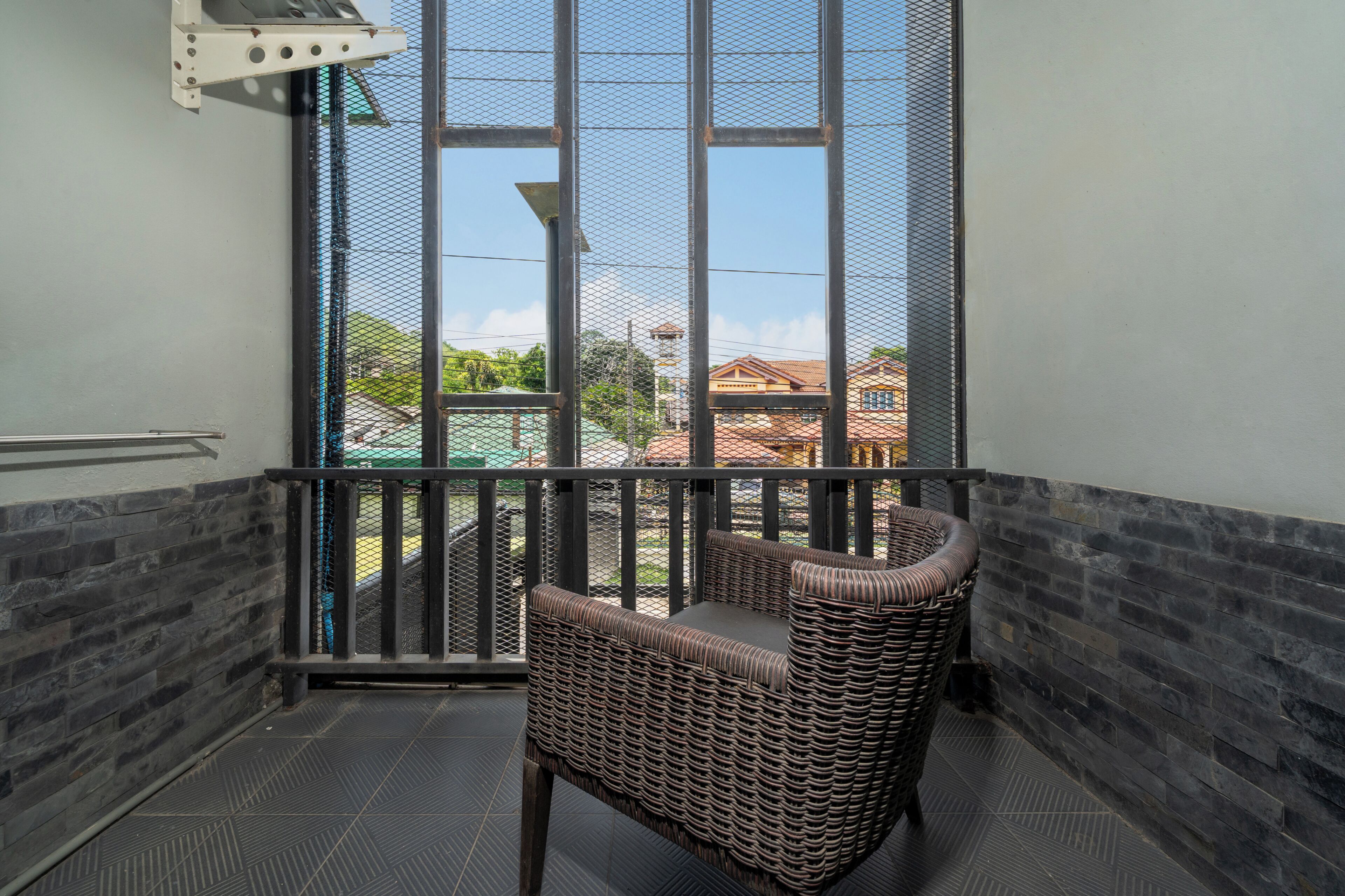 Uma varanda com cadeira de vime, guarda-corpo de metal e vista para edifícios e vegetação.