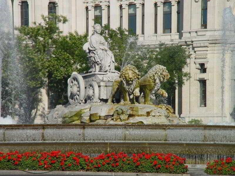 Uma fonte clássica com estátua de mulher e leões, rodeada por vegetação e um edifício com colunas ao fundo.