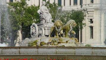 Uma fonte clássica com estátua de mulher e leões, rodeada por vegetação e um edifício com colunas ao fundo.