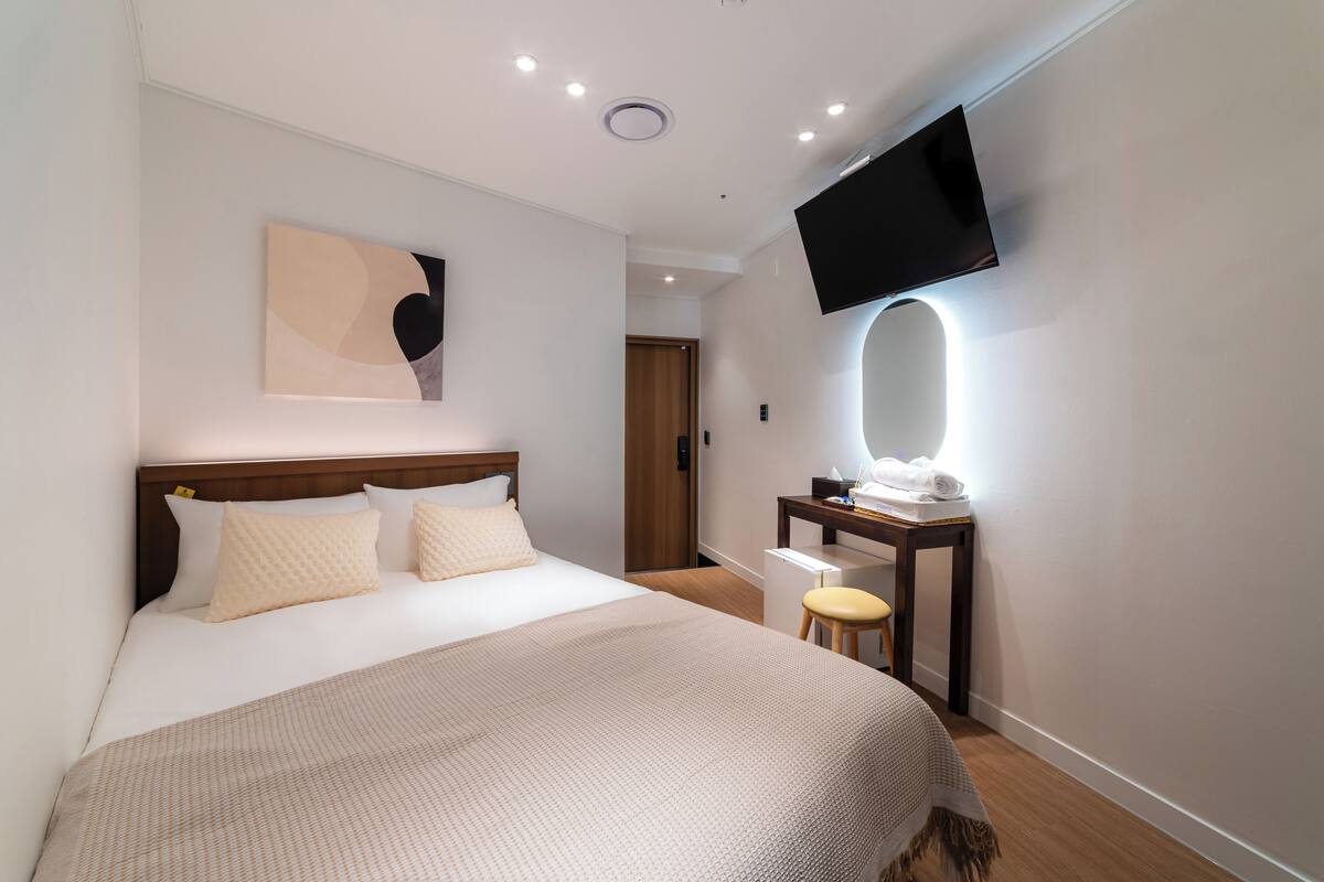 Quarto de hotel com cama, escrivaninha com espelho, televisão na parede e porta que dá para outro cômodo.