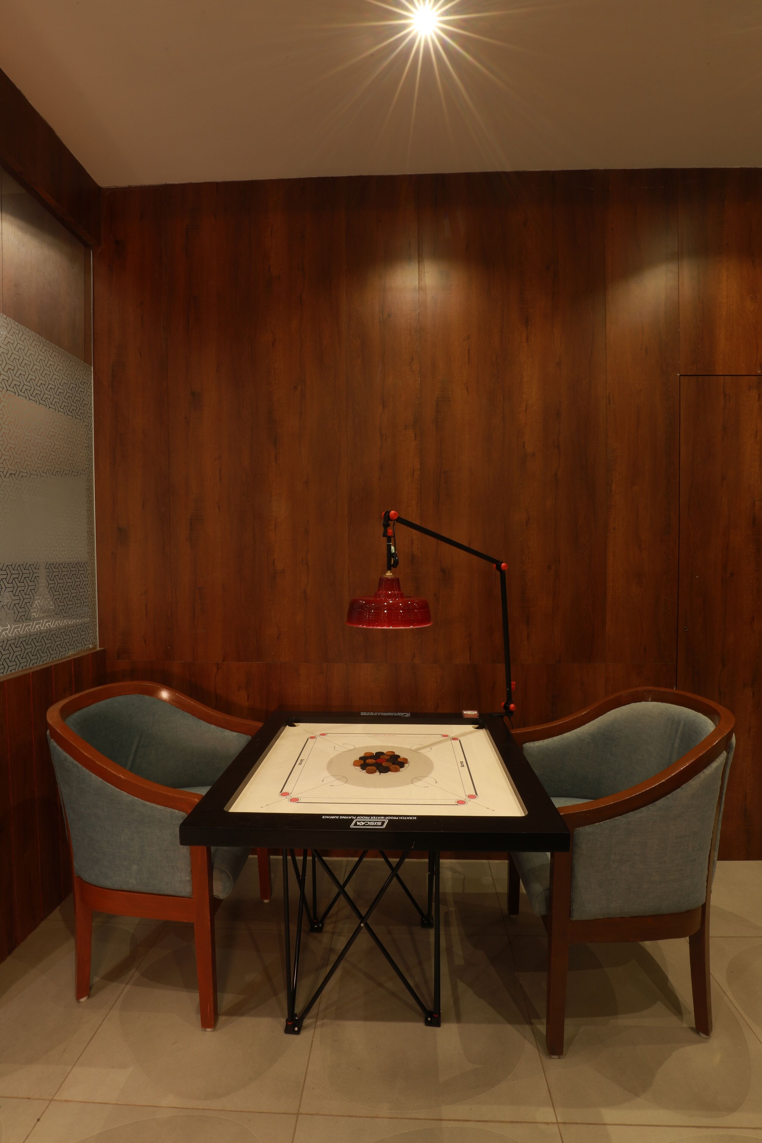 Quarto com paredes de madeira, uma mesa com abajur vermelho e duas cadeiras com estofamento azul claro.