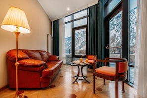 Quarto com sofá de couro, uma cadeira de madeira, mesa com bandeja e amplas janelas com vista para as montanhas.