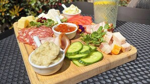Uma tábua de madeira com diversos alimentos, uma bebida com canudo e um muro de tijolos ao fundo.