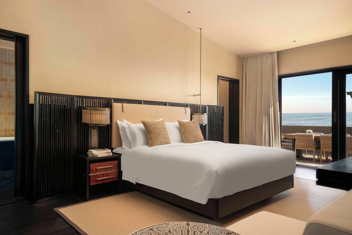 Quarto de hotel com uma cama grande, mesa de cabeceira, luminária, vista para o mar e varanda com mesa e cadeiras.