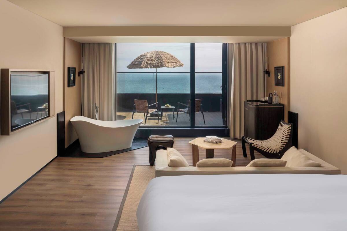 Quarto de hotel com uma cama grande, uma banheira, varanda com mesa e cadeiras, e vista para o mar.