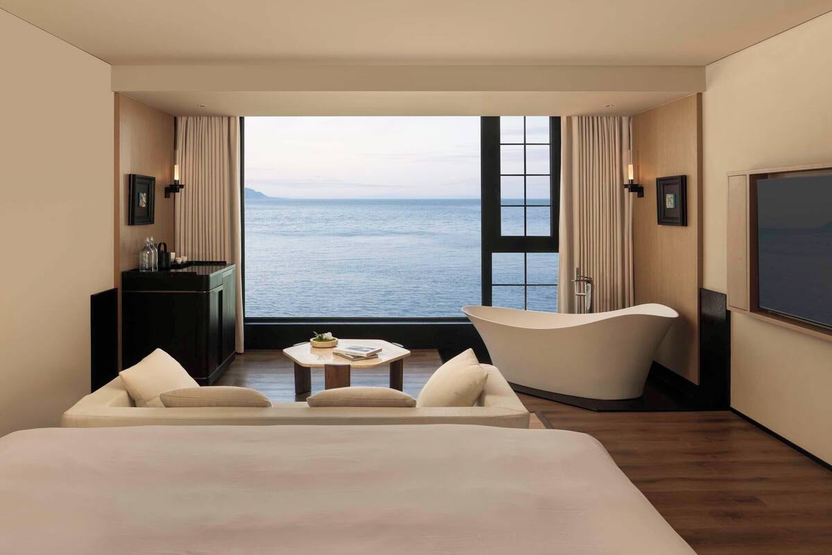 Quarto de hotel com uma cama, área de estar com mesa, banheira e uma grande janela com vista para o mar.