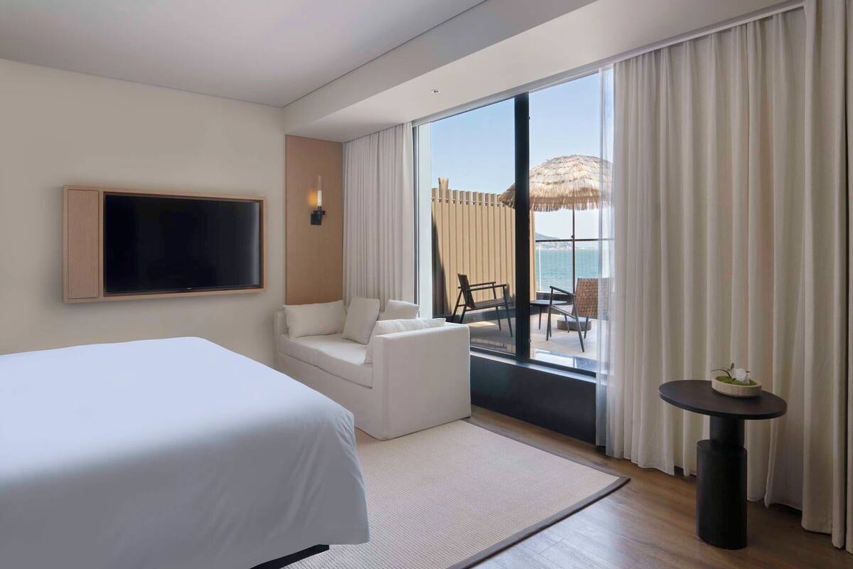 Quarto de hotel com cama, sofá, mesa pequena, TV e varanda com vista para o mar.