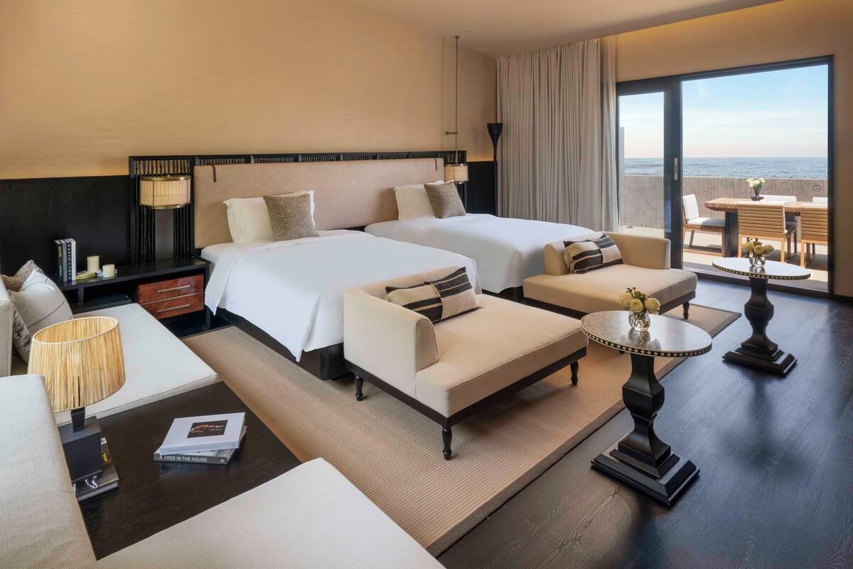 Quarto de hotel com duas camas, um sofá, uma mesa de centro, uma luminária e vista para o mar.