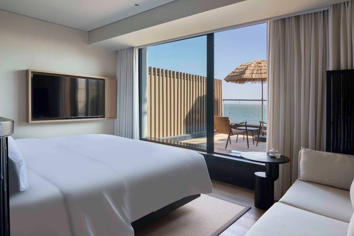 Quarto de hotel com uma cama grande, televisão, varanda com vista para o mar e área de estar com mesa e cadeiras.