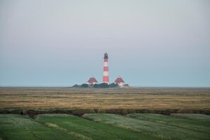 Um farol com torre listrada em vermelho e branco, dois edifícios menores com telhados vermelhos e uma paisagem plana.