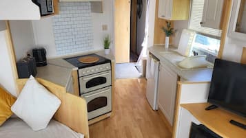 Uma kitchenette compacta com fogão, forno e pia, uma pequena área de estar com sofá e uma televisão sobre uma mesa de madeira.