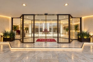 Entrada de hotel com porta de vidro, tapete vermelho com o nome do hotel e plantas em vasos de cada lado.