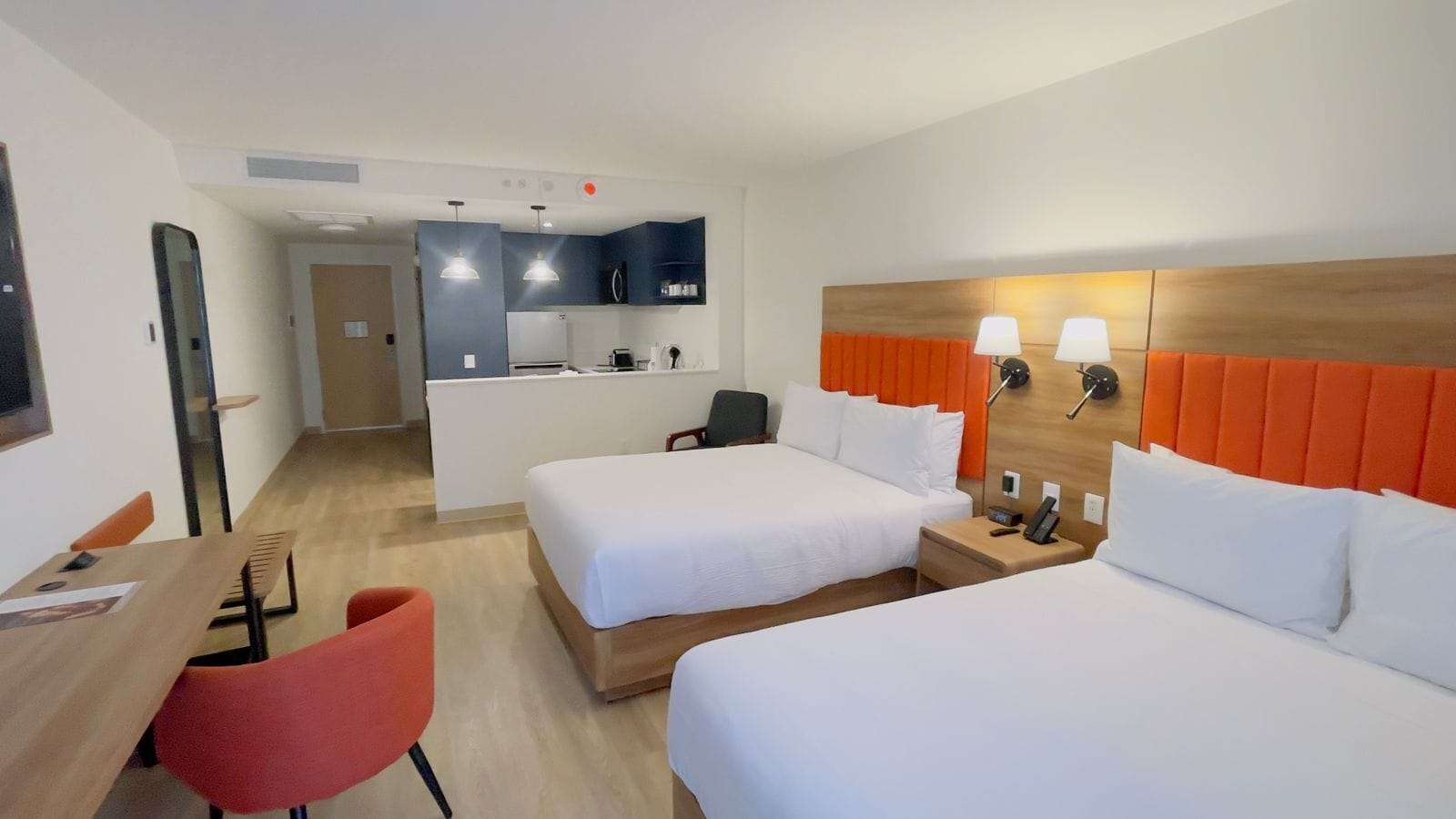 Quarto de hotel com duas camas, uma escrivaninha, uma cadeira, uma kitchenette e um banheiro.