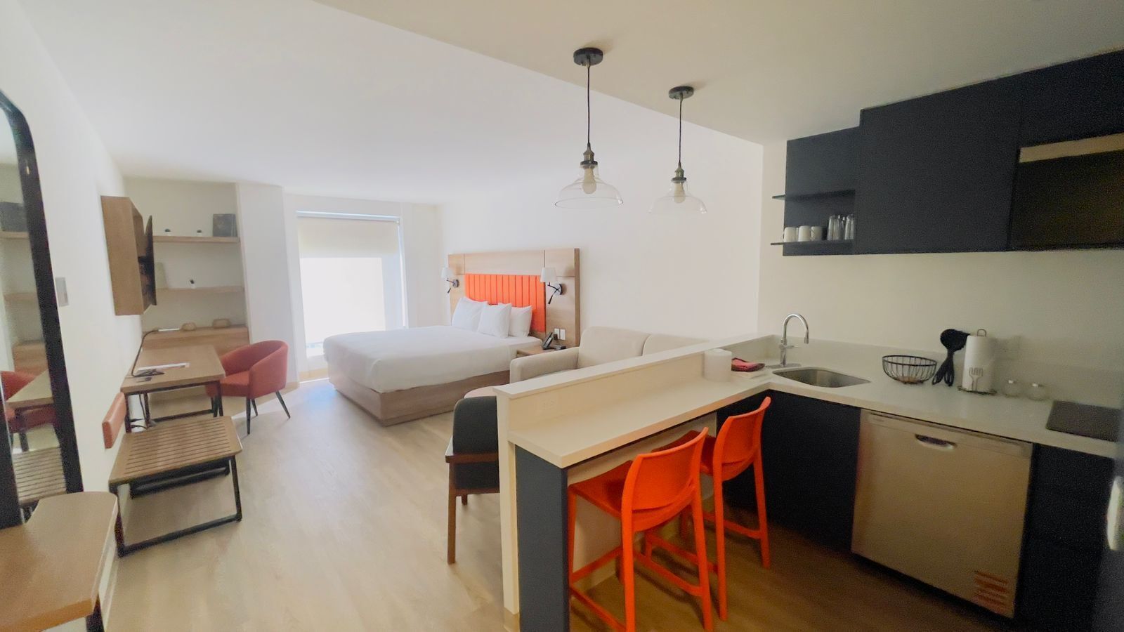 Quarto de hotel moderno com cozinha compacta, cama, escrivaninha e área de refeições.