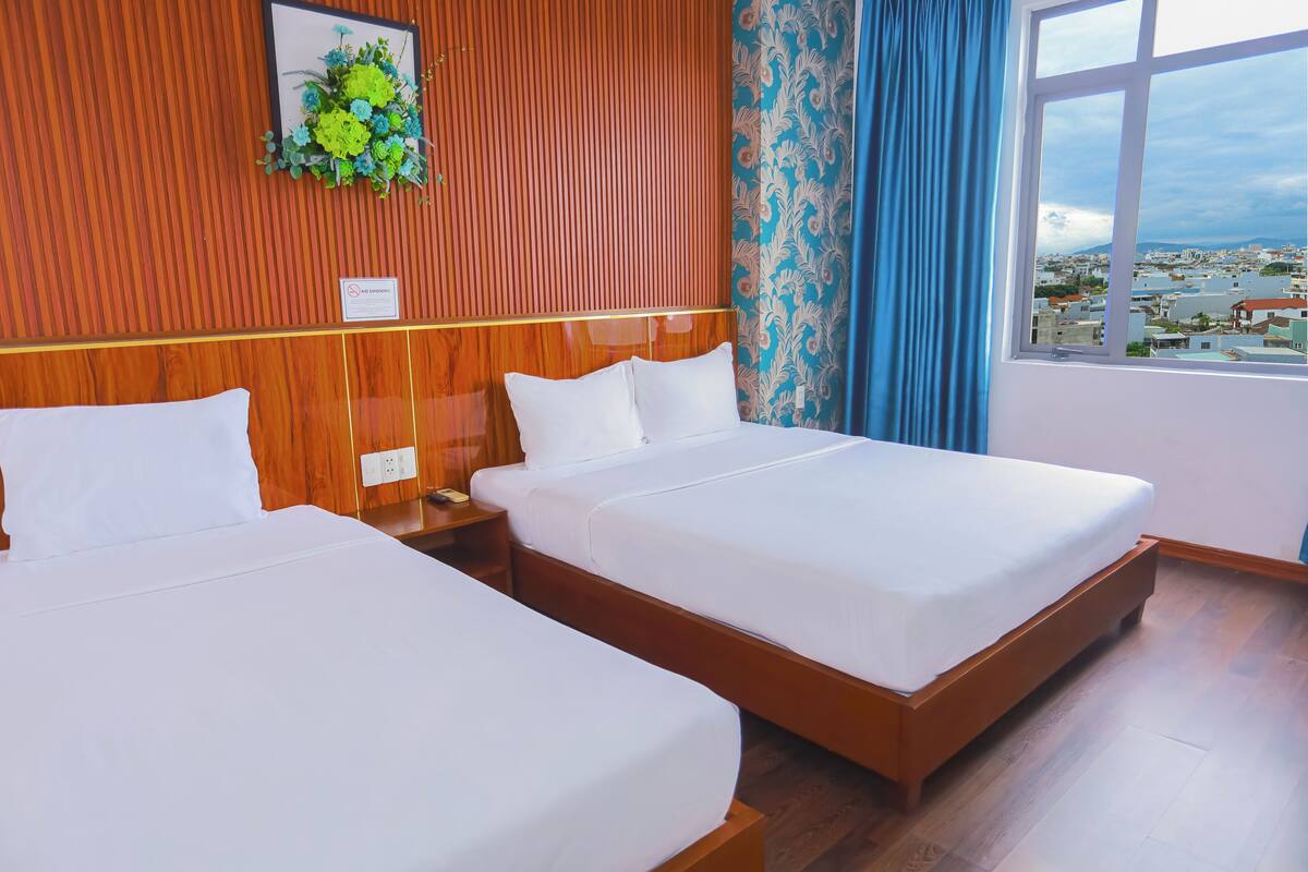 Quarto de hotel com duas camas, cabeceira de madeira, janela com vista e um arranjo floral fixado na parede.