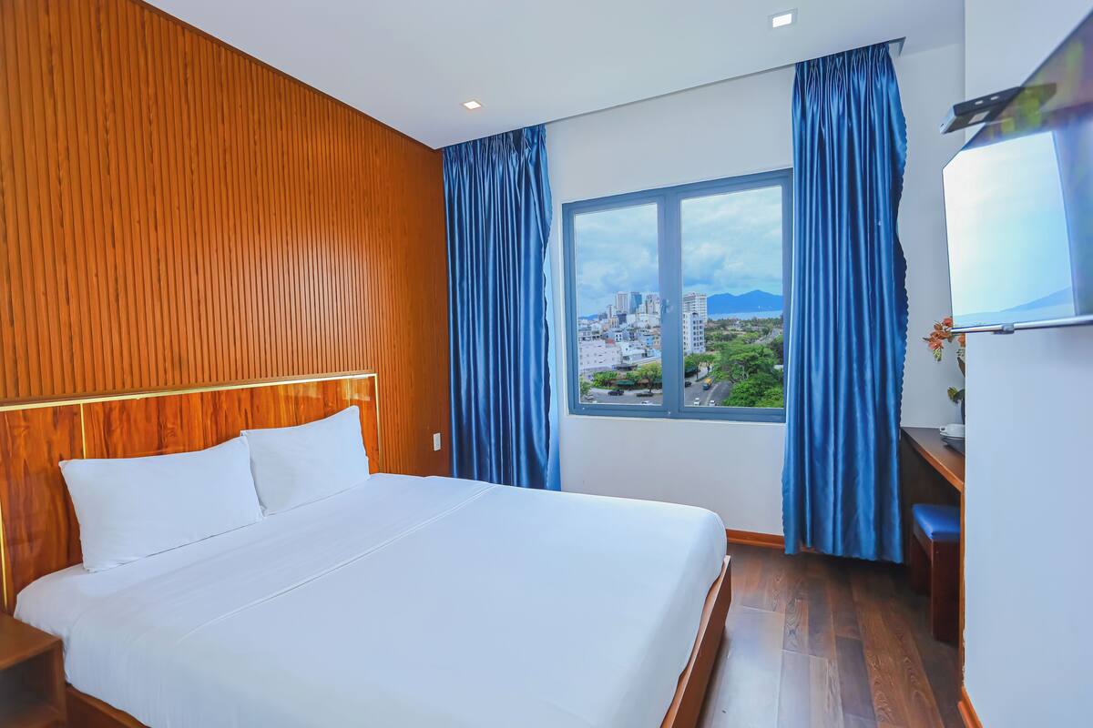 Quarto de hotel com uma cama grande, painéis de madeira na parede, uma mesa e uma janela com cortinas.