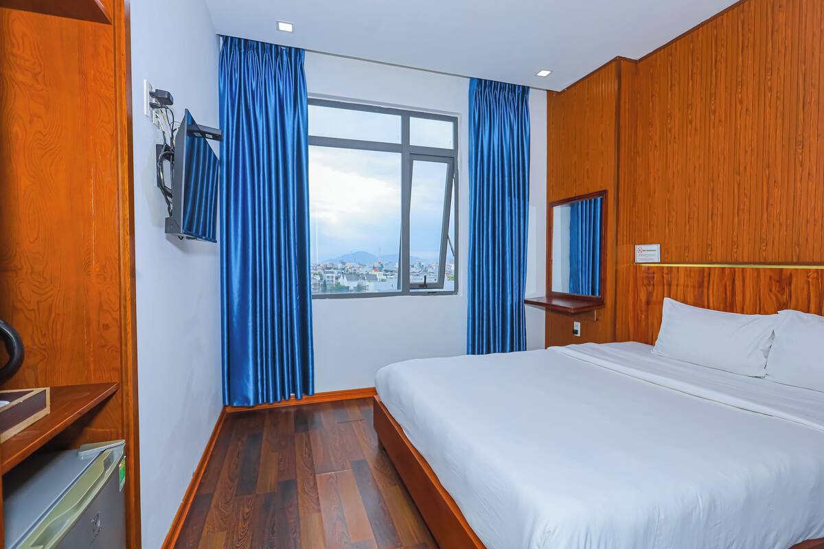 Quarto de hotel com uma cama grande, painéis de madeira, uma janela com cortinas, uma TV de tela plana e vista para a cidade.