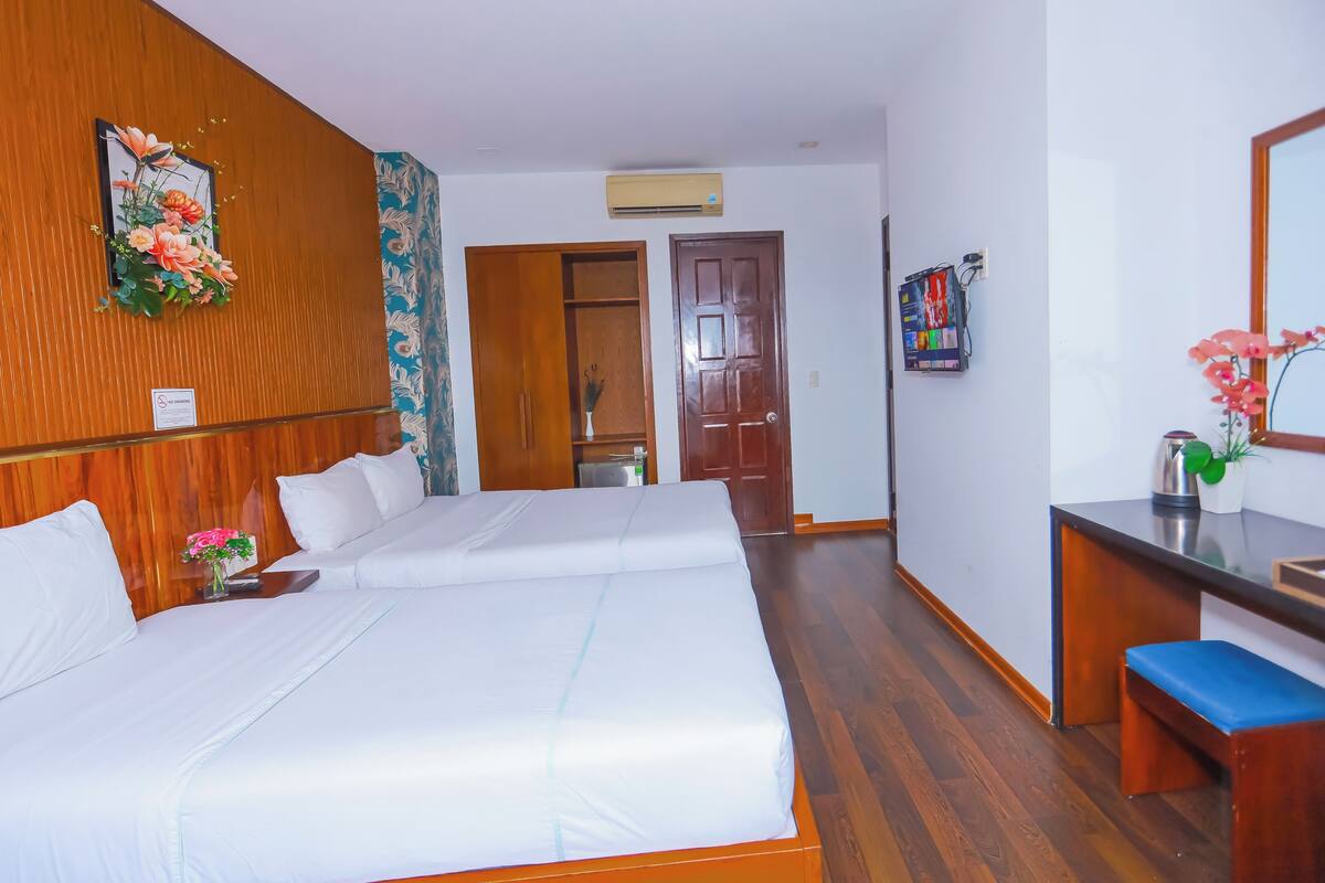 Quarto de hotel com duas camas, um guarda-roupa de madeira, uma escrivaninha com espelho e um pequeno banco.