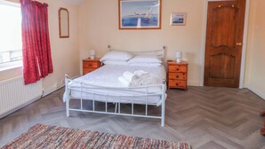 Quarto com cama, duas mesas de cabeceira, porta de madeira, radiador e janela com cortinas vermelhas.