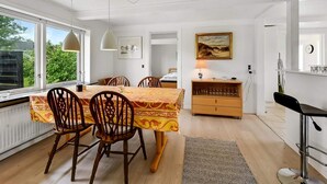 Uma sala de jantar com mesa, cadeiras, luminárias pendentes, um armário de madeira e vista através de grandes janelas.