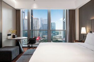 Quarto de hotel com uma cama grande, uma mesa pequena com um vaso, uma cadeira, uma mesa de cabeceira com uma luminária e vista para edifícios da cidade através de janelas amplas.