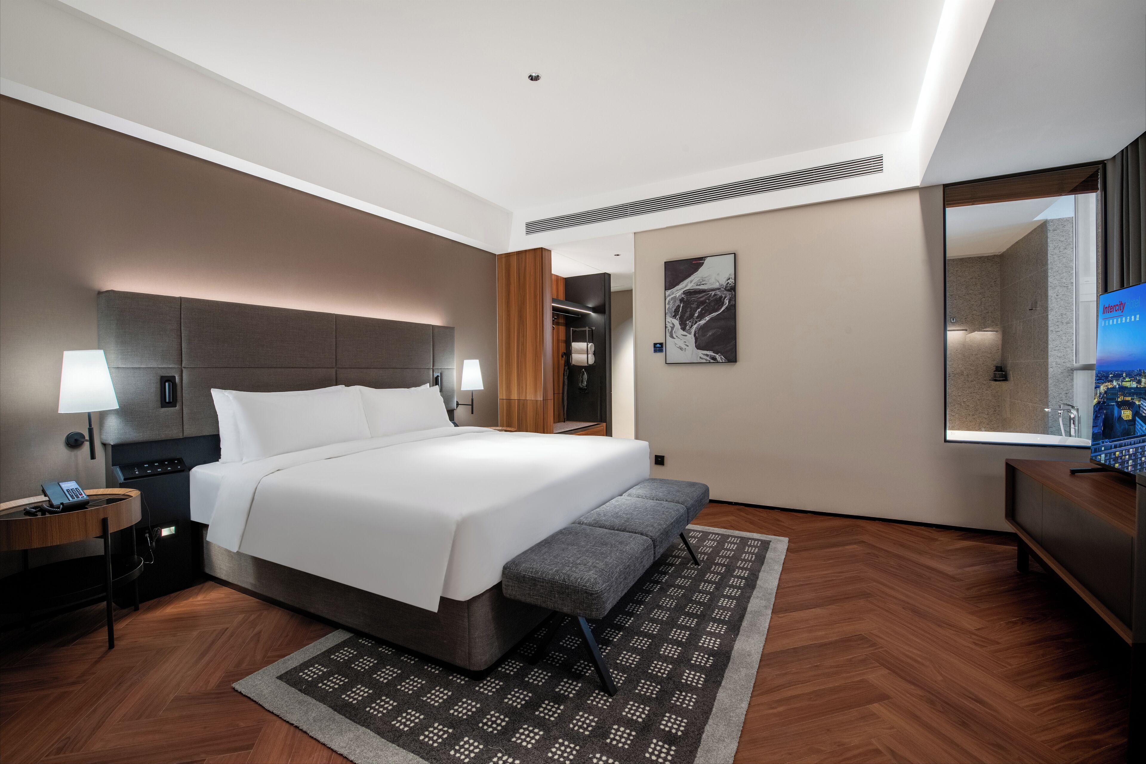 Quarto de hotel moderno com cama grande, mesas de cabeceira, um banco, escrivaninha, espelho e televisão presa à parede.