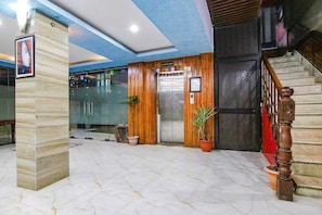 Um hall com piso de mármore, painéis de madeira, escadaria e elevador de vidro.
