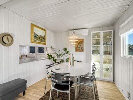 Uma área de refeições com uma mesa branca, cadeiras pretas, um banco cinzento, uma planta em vaso e um armário com louças.