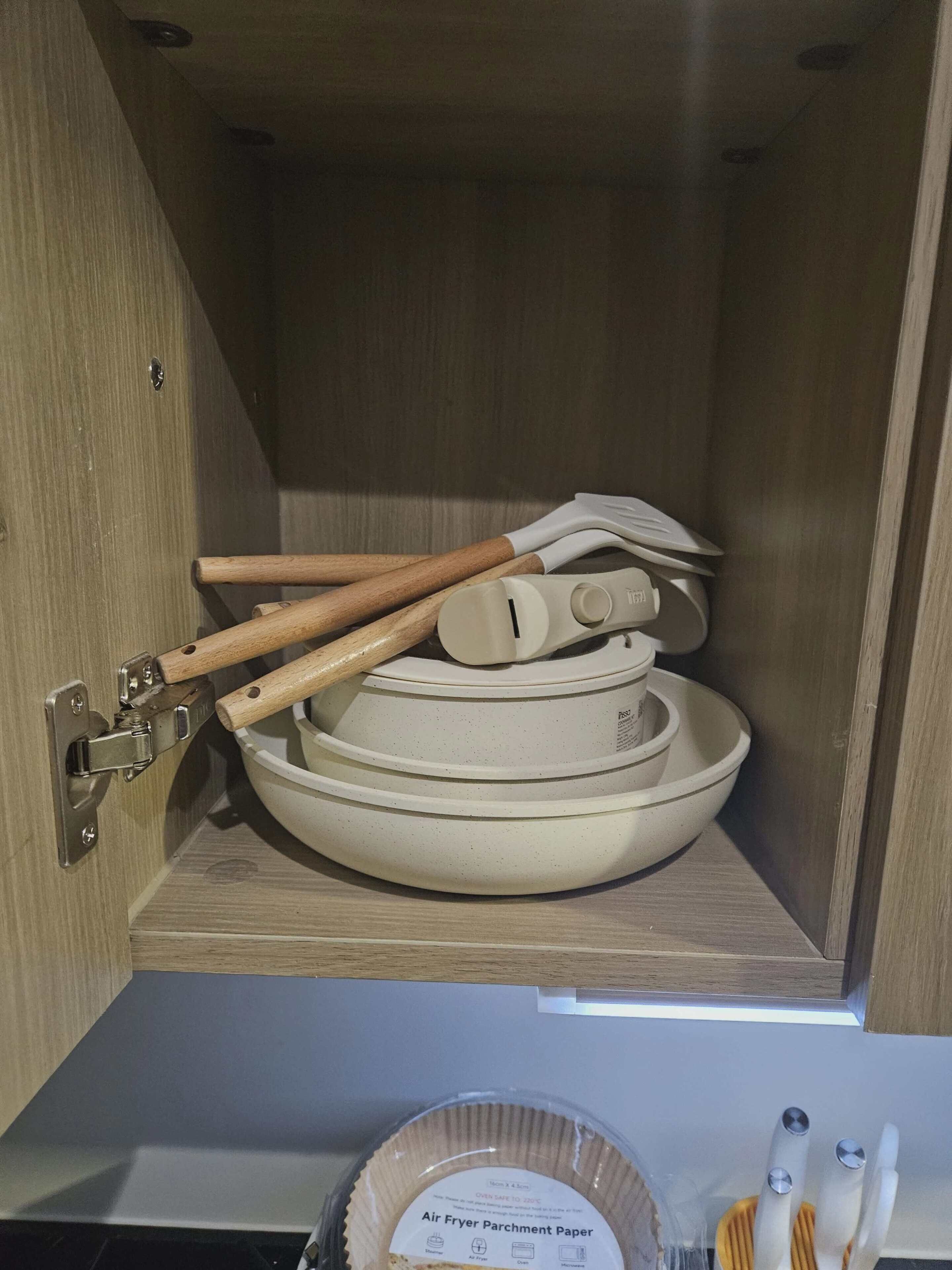 Um armário de cozinha com interior de madeira, contendo uma pilha de tigelas brancas, uma tigela de mistura branca e utensílios de madeira.